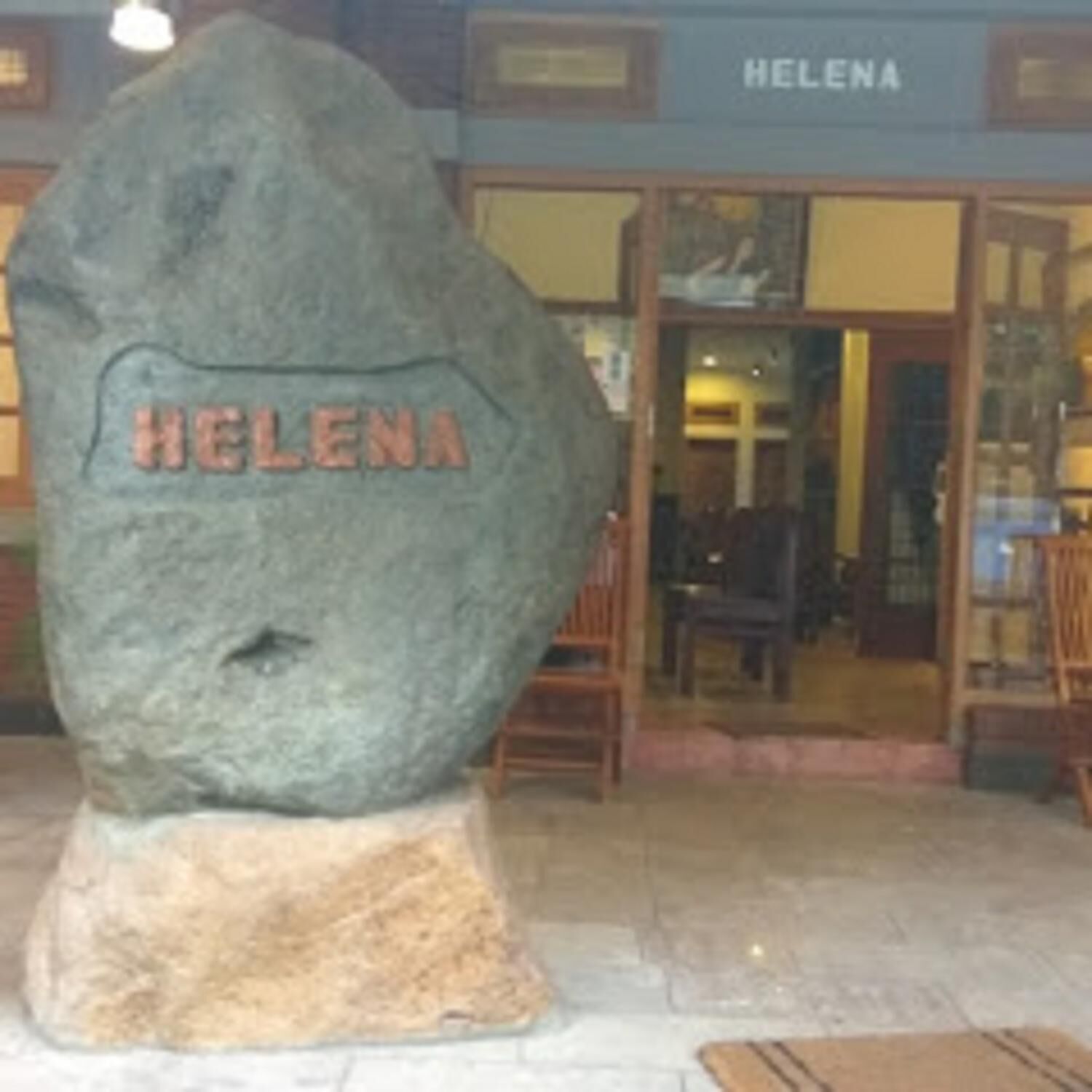 Uma grande escultura em pedra com o nome "HELENA" gravado, colocada em frente a um edifício com entrada de vidro.