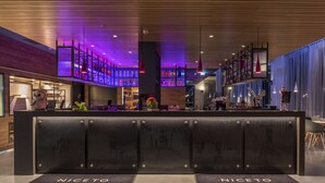 Uma área de recepção de hotel com design moderno, com balcão de recepção, espaço de bar com prateleiras de garrafas e uma grande escultura decorativa.