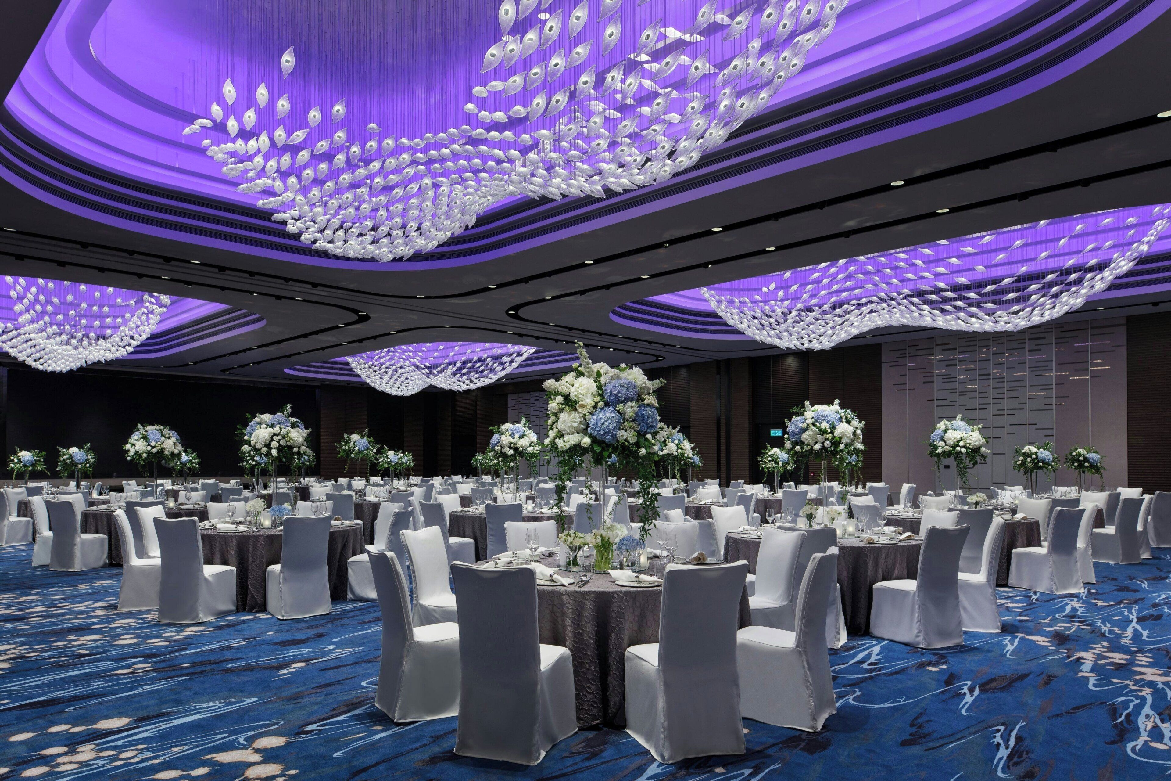 Salão de banquetes com mesas redondas, cadeiras brancas e iluminação roxa ambiente.