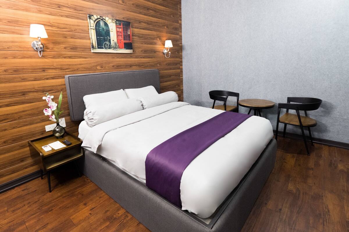 Quarto de hotel moderno com parede de madeira, uma cama grande com roupa de cama branca e roxa, duas luminárias de cabeceira, uma mesinha com um vaso de flores e duas cadeiras perto de uma mesa redonda.