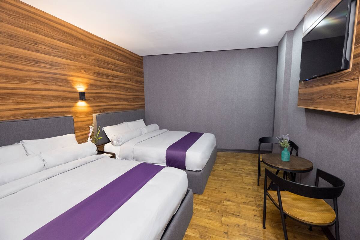 Quarto de hotel com duas camas, uma mesa pequena e uma televisão fixada na parede.