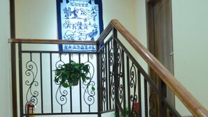 Escadaria com grades de metal ornamentadas e corrimão de madeira, que leva a um patamar com um cartaz emoldurado e um vaso de planta.