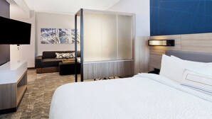 Quarto de hotel moderno com cama grande, sofá, televisão e luminária fixada na parede.