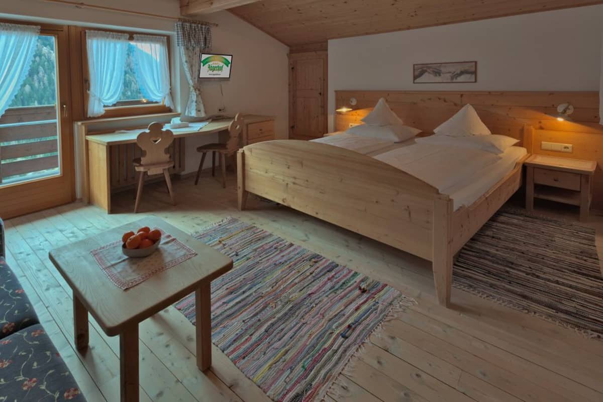 Quarto aconchegante com uma cama de madeira, uma escrivaninha e uma cadeira. Há uma pequena mesa com uma tigela de frutas.