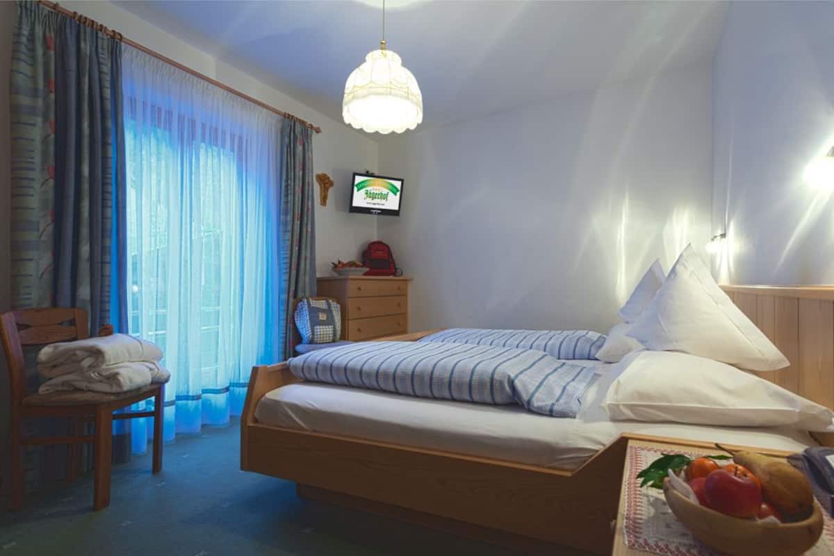 Quarto de hotel com cama, poltrona, escrivaninha com luminária, televisão e janela com cortinas.