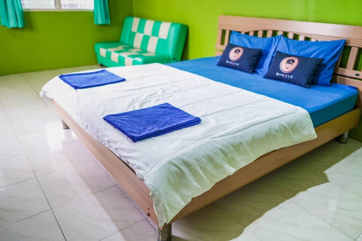 Uma cama com colcha branca e azul, duas almofadas azuis e um sofá verde xadrez em um quarto com paredes verdes e piso de azulejos brancos.