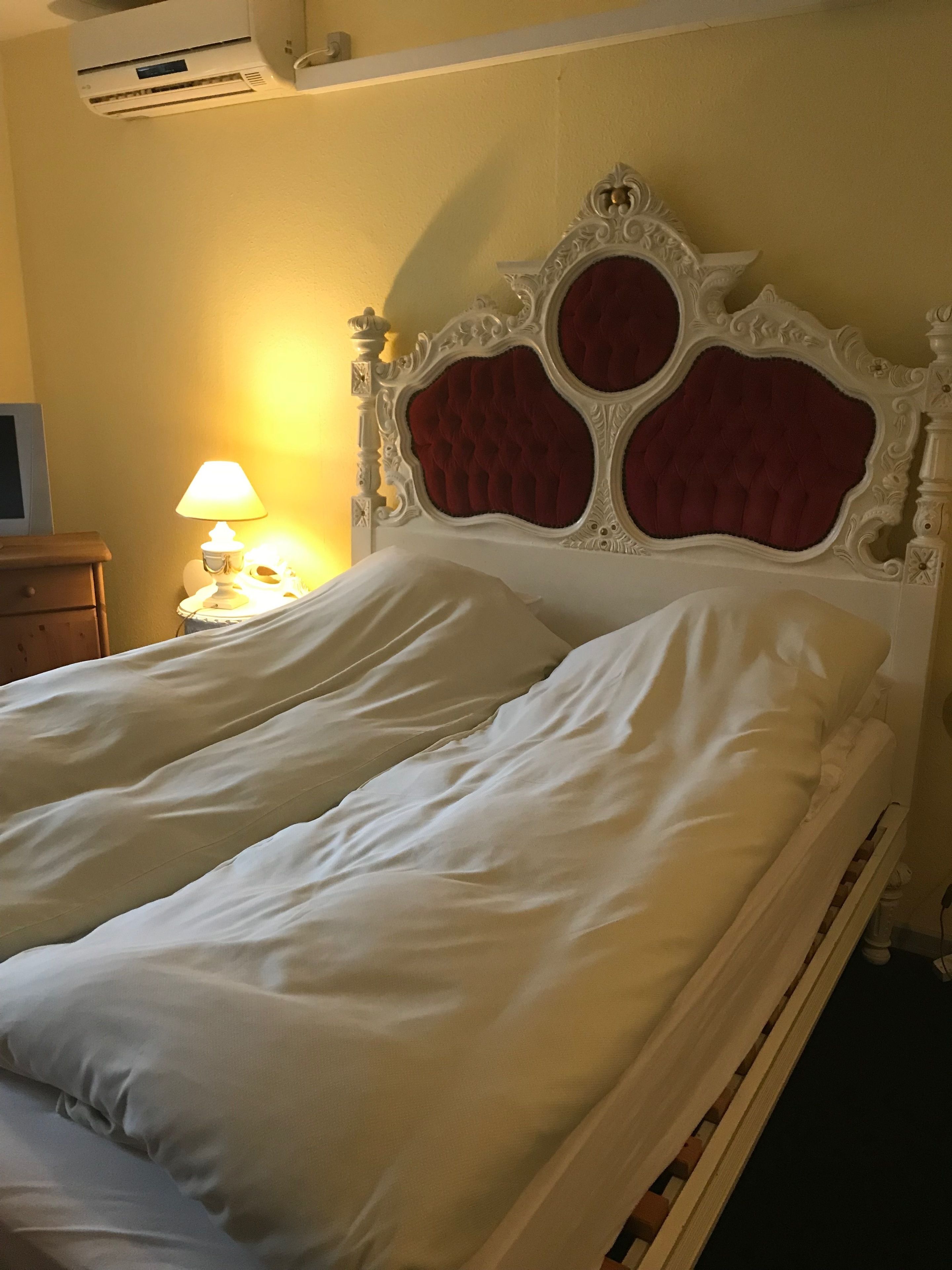 Uma cama de casal com estrutura branca e cabeceira vermelha, uma luminária de cabeceira e um aparelho de ar condicionado na parede.