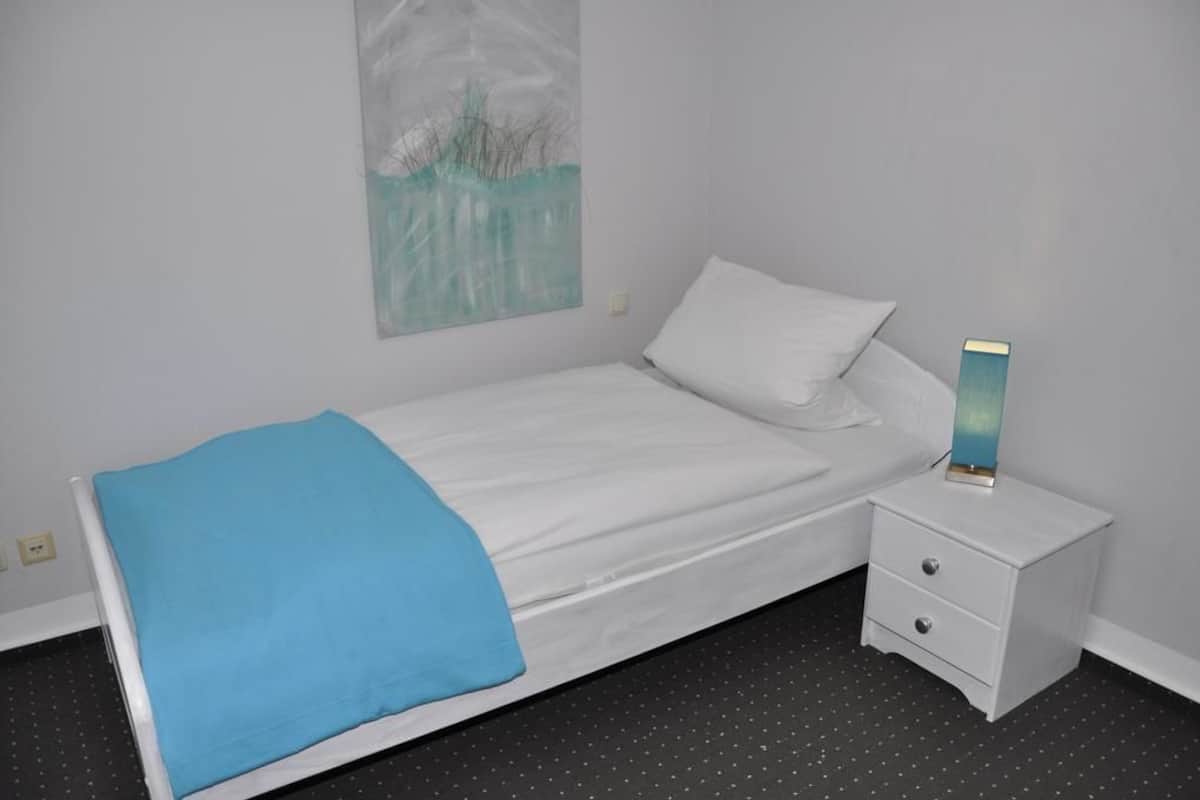 Uma cama de solteiro com colchão branco e cobertor azul, uma mesinha de cabeceira branca com gavetas e um quadro abstrato pendurado na parede.