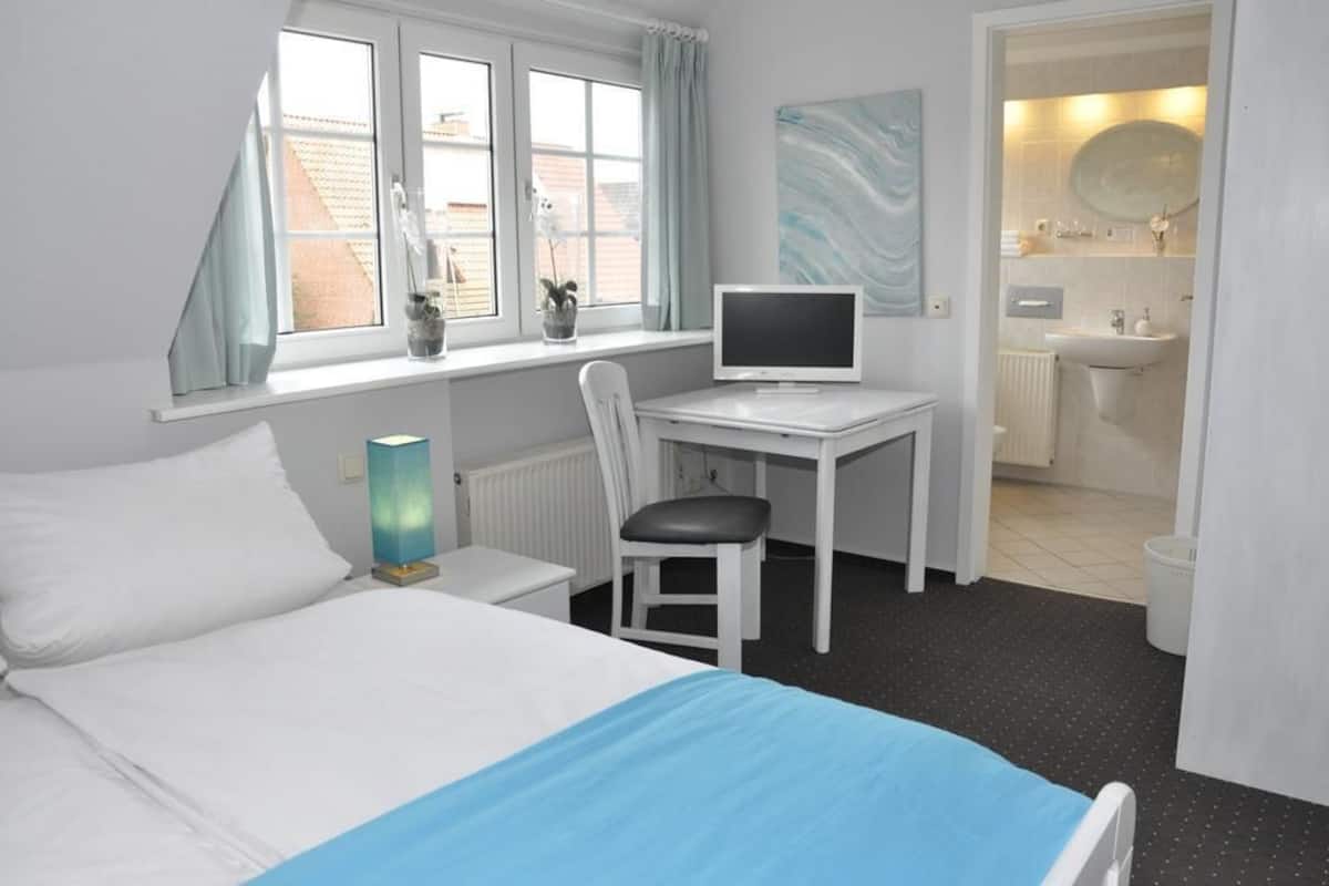 Quarto com cama, escrivaninha com computador, cadeira, janela com cortinas e banheiro visível através de uma porta aberta.