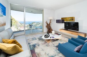 Uma sala de estar moderna com um sofá, uma poltrona azul, uma mesa de centro, uma televisão e vista para o mar através de grandes portas de vidro.