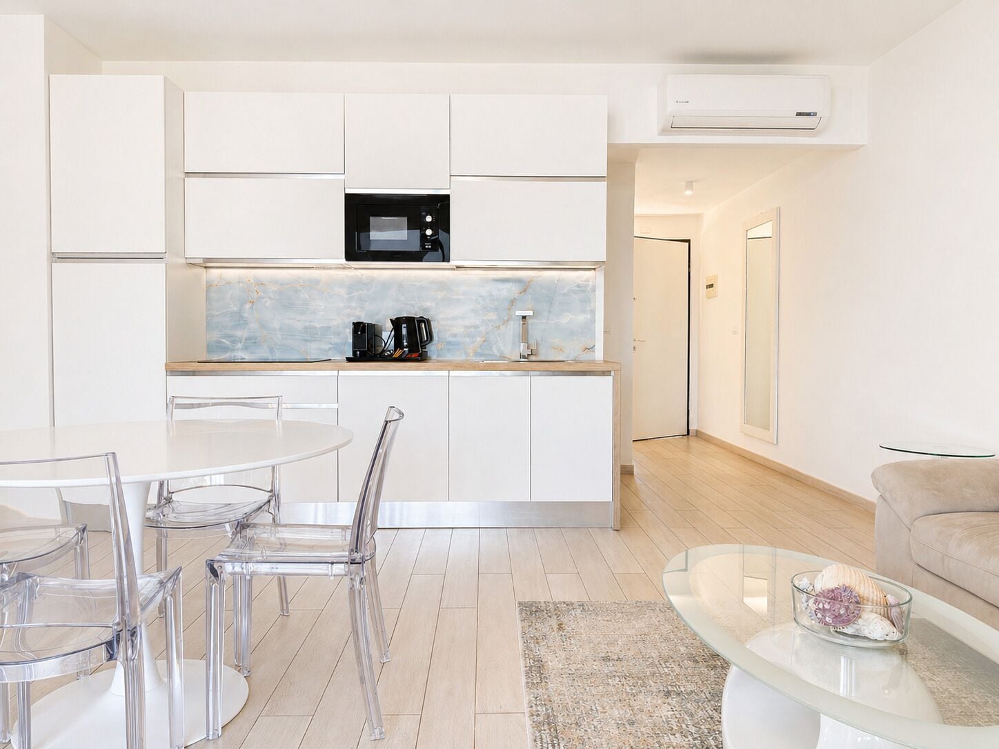 Uma cozinha moderna com armários brancos, micro-ondas e cafeteira. Há uma mesa de jantar com cadeiras transparentes e uma mesa de centro de vidro com objetos decorativos.
