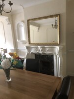 Uma sala de jantar com mesa de madeira, cadeiras, um vaso com flores, uma lareira branca com espelho dourado em cima e um lustre.