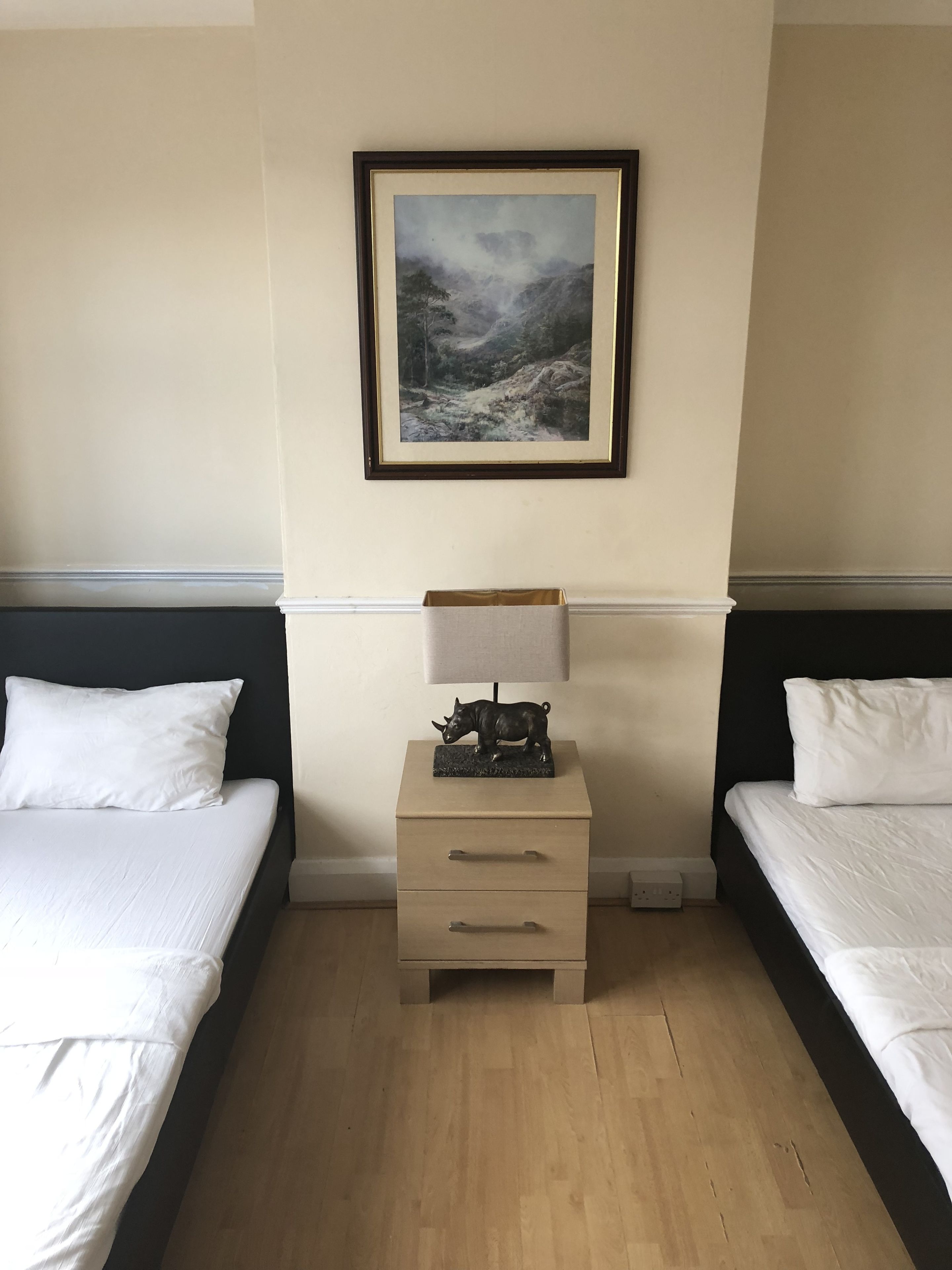 Quarto de hotel com duas camas, uma mesa de cabeceira com abajur e um quadro na parede.