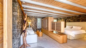 Quarto com parede de pedra, vigas de madeira no teto, cama, sofá branco e mesa de cabeceira de madeira.