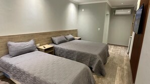 Quarto de hotel com duas camas, uma cabeceira de madeira, uma mesa de cabeceira, uma televisão e um ar condicionado.