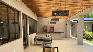 Uma área de pátio externa com teto de madeira, assentos acolchoados e um espaço de bar com refrigeradores.