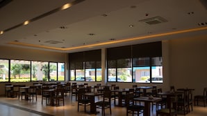 Um espaço amplo e interno, com várias mesas e cadeiras dispostas em fileiras, grandes janelas oferecendo vistas para o exterior e iluminação no teto.