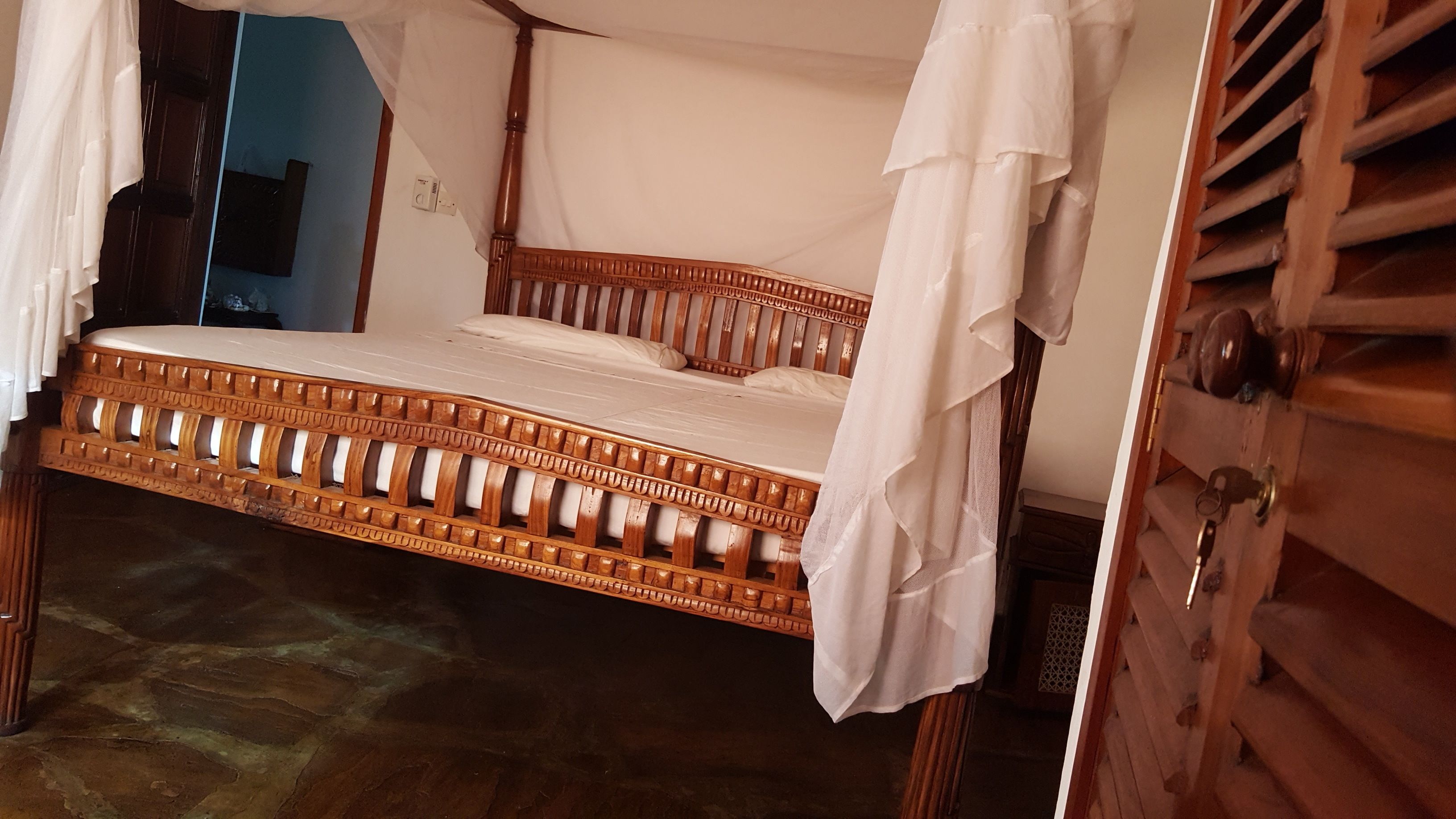 Uma cama com estrutura de madeira e roupa de cama branca, uma porta de madeira com fechadura e uma tomada elétrica na parede.