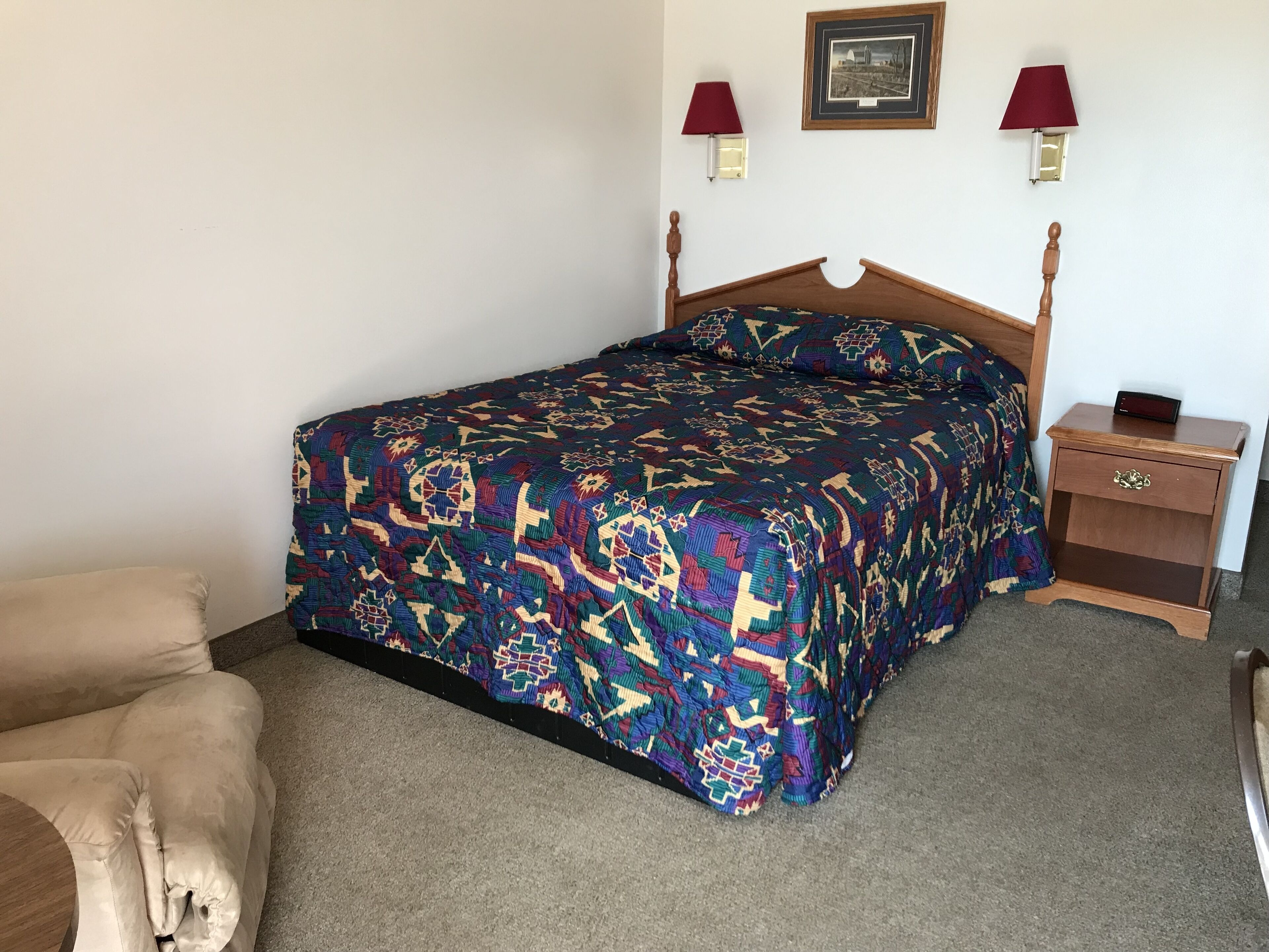 Cama bem arrumada com colcha estampada, mesa de cabeceira de madeira com luminária e uma cadeira em quarto com paredes lisas.