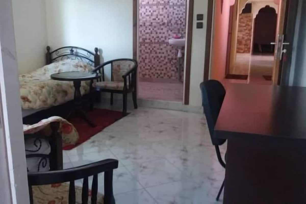 Quarto pequeno e compacto com uma cama, uma mesa e duas cadeiras. Há um tapete vermelho no chão e as paredes são revestidas de azulejos.