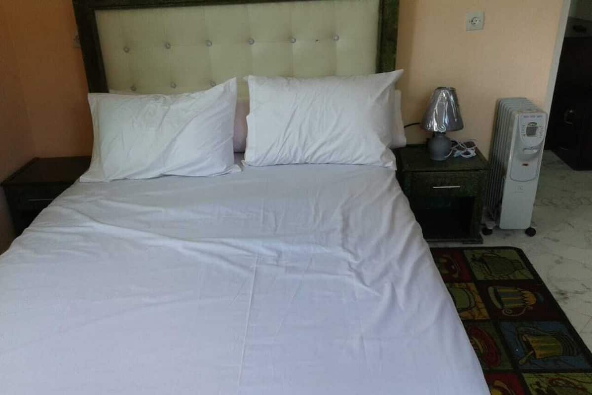 Cama bem arrumada com lençóis brancos, cabeceira estofada e mesa de cabeceira com abajur e aquecedor.
