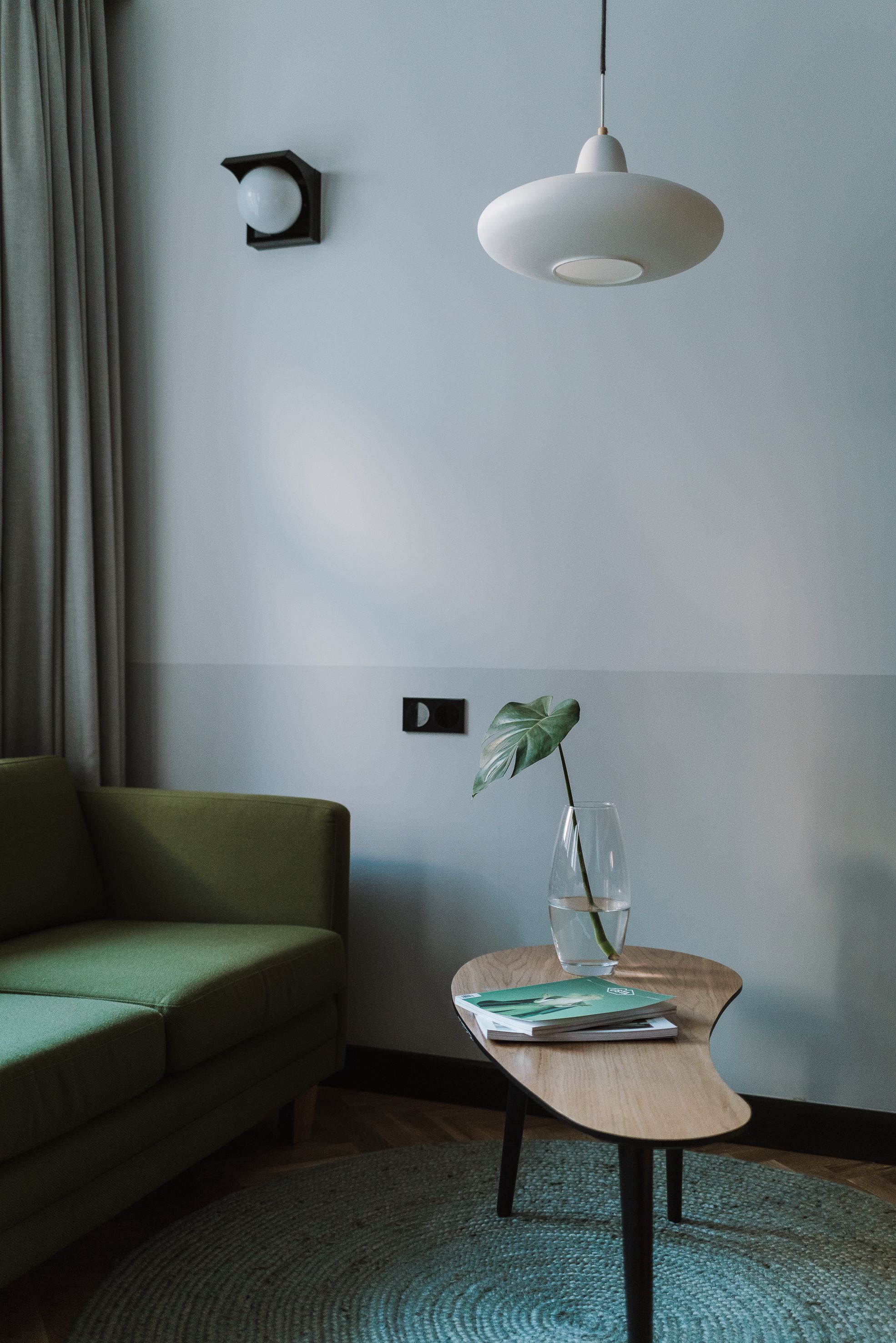 Sala de estar moderna com um sofá verde, uma mesa de centro de madeira com uma planta e um livro, uma luminária pendente branca e um aplique de parede.