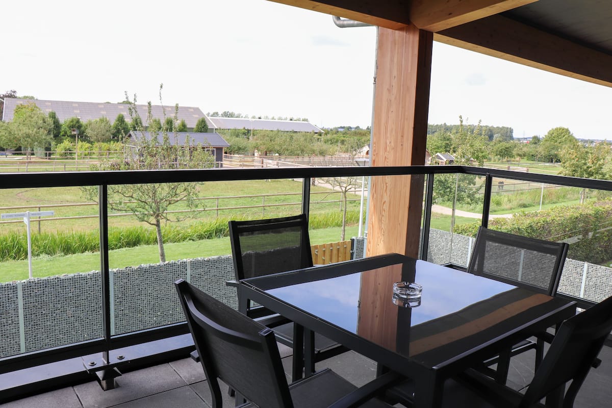 Uma varanda com mesa e cadeiras de vidro, proporcionando vista para uma paisagem verde e edifícios.