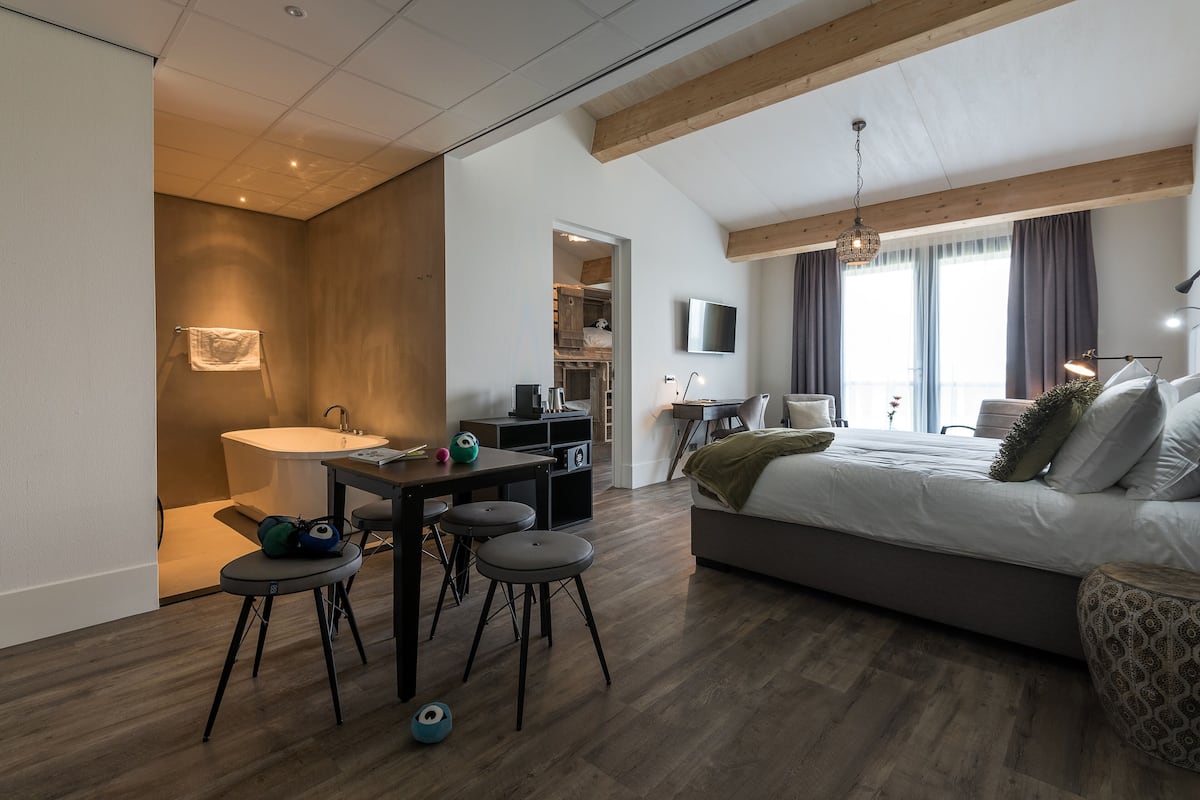 Quarto moderno com uma cama grande, uma escrivaninha com bancos, um banheiro pequeno e uma TV fixada na parede.