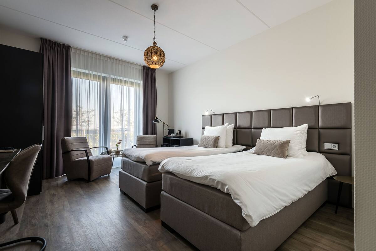 Quarto de hotel moderno com duas camas, uma escrivaninha, uma cadeira e uma janela grande.