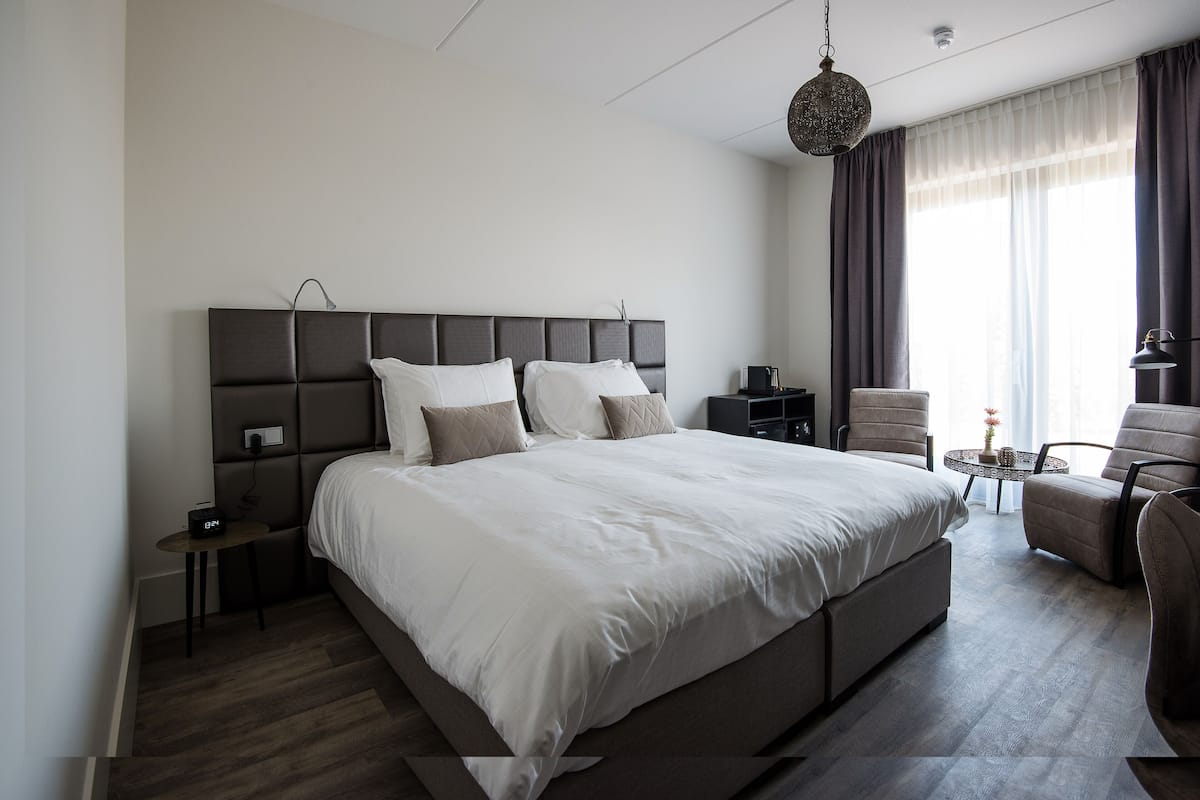 Quarto de hotel moderno com uma cama grande, uma mesinha de cabeceira, uma cadeira, uma pequena mesa redonda e um lustre.