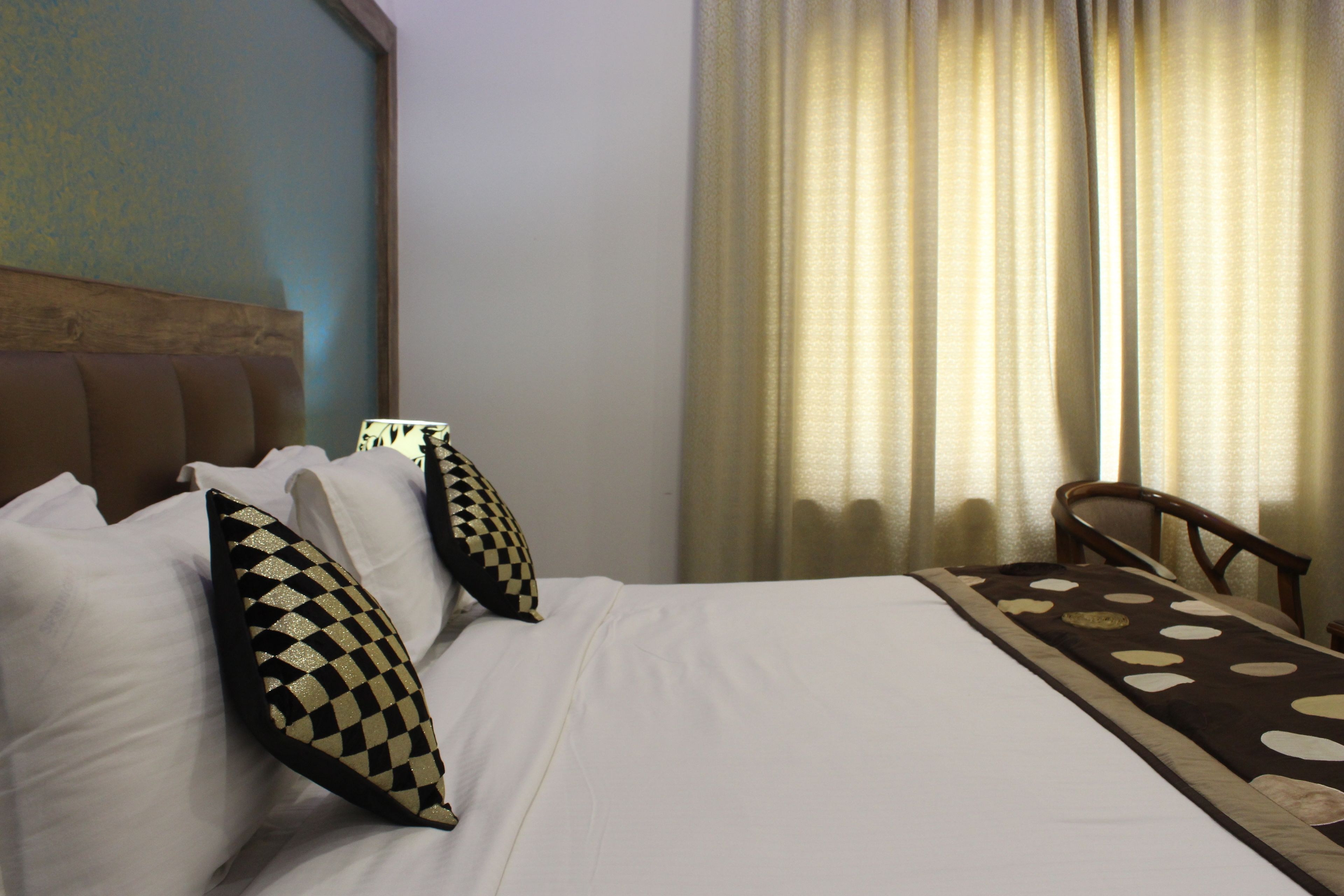 Uma cama bem arrumada com lençóis brancos e almofadas estampadas, cabeceira de madeira e vista para uma janela com cortinas.