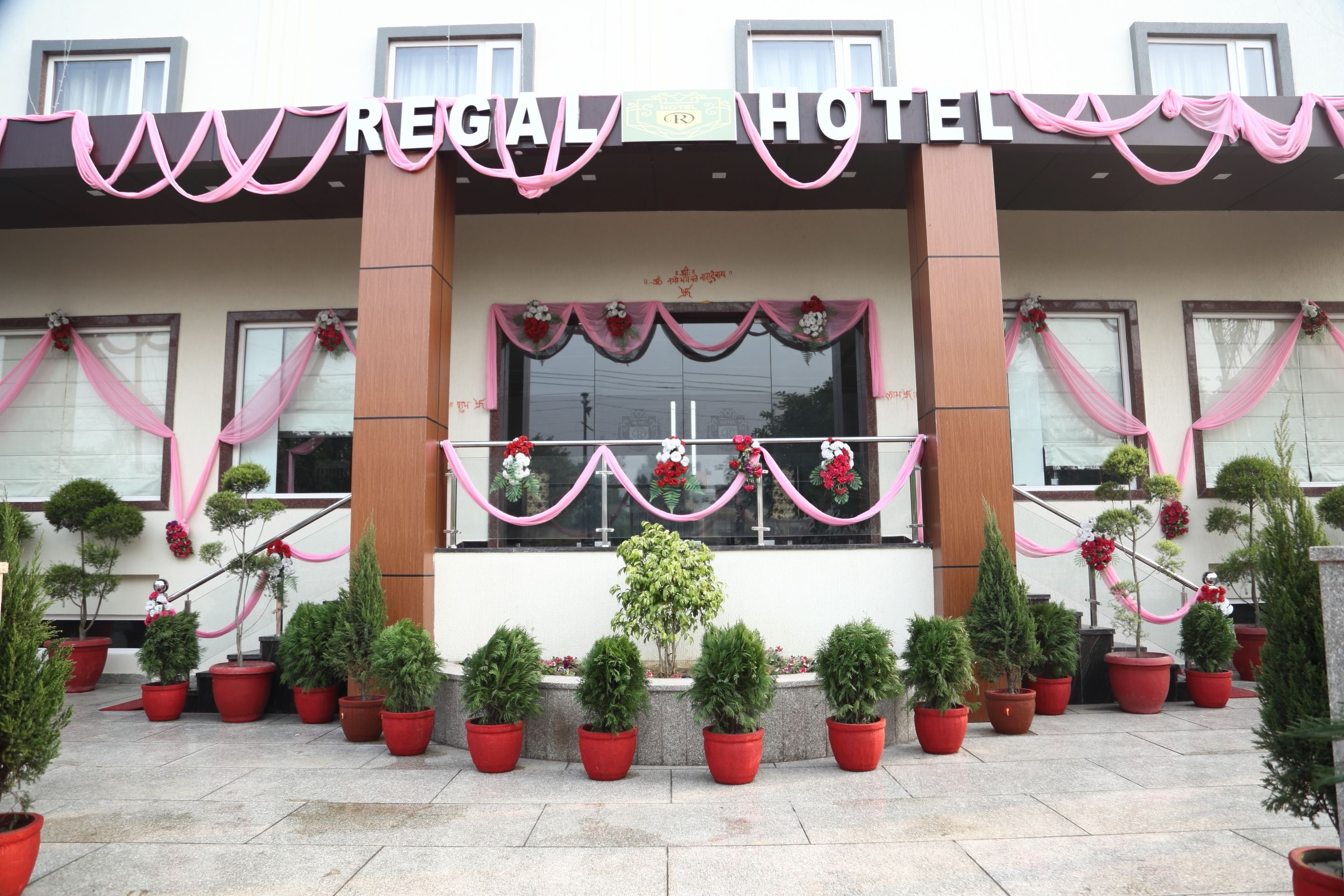 Entrada de um hotel com uma placa indicando "Regal Hotel", rodeada por plantas em vasos e cortinas cor-de-rosa.