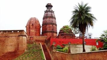 Uma fortaleza histórica com uma grande torre central e uma cúpula menor, cercada por uma muralha fortificada e uma palmeira.