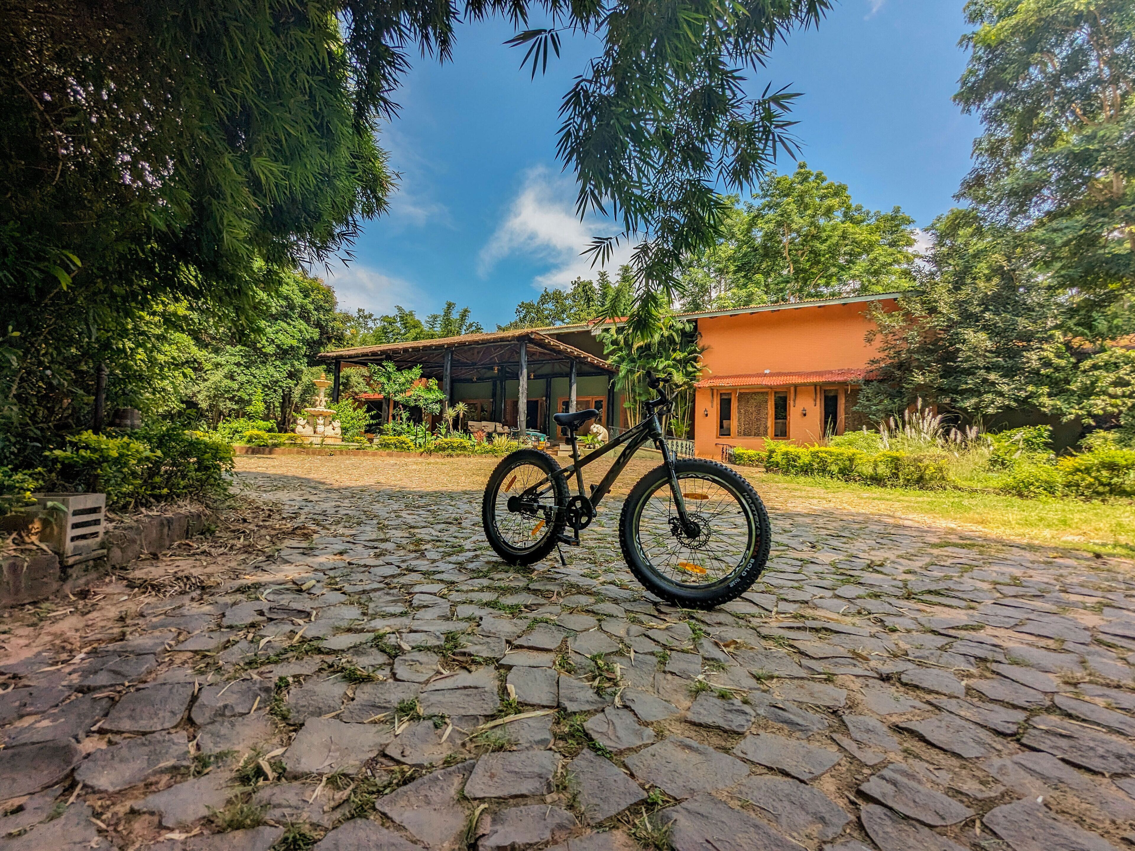 Uma bicicleta estacionada em um caminho de pedras irregulares, em frente a um edifício com telhado de azulejos e uma fonte ao fundo.