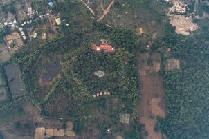 Vista aérea de uma propriedade com um edifício central rodeado por vegetação densa, um espelho d'água circular e algumas estruturas menores.