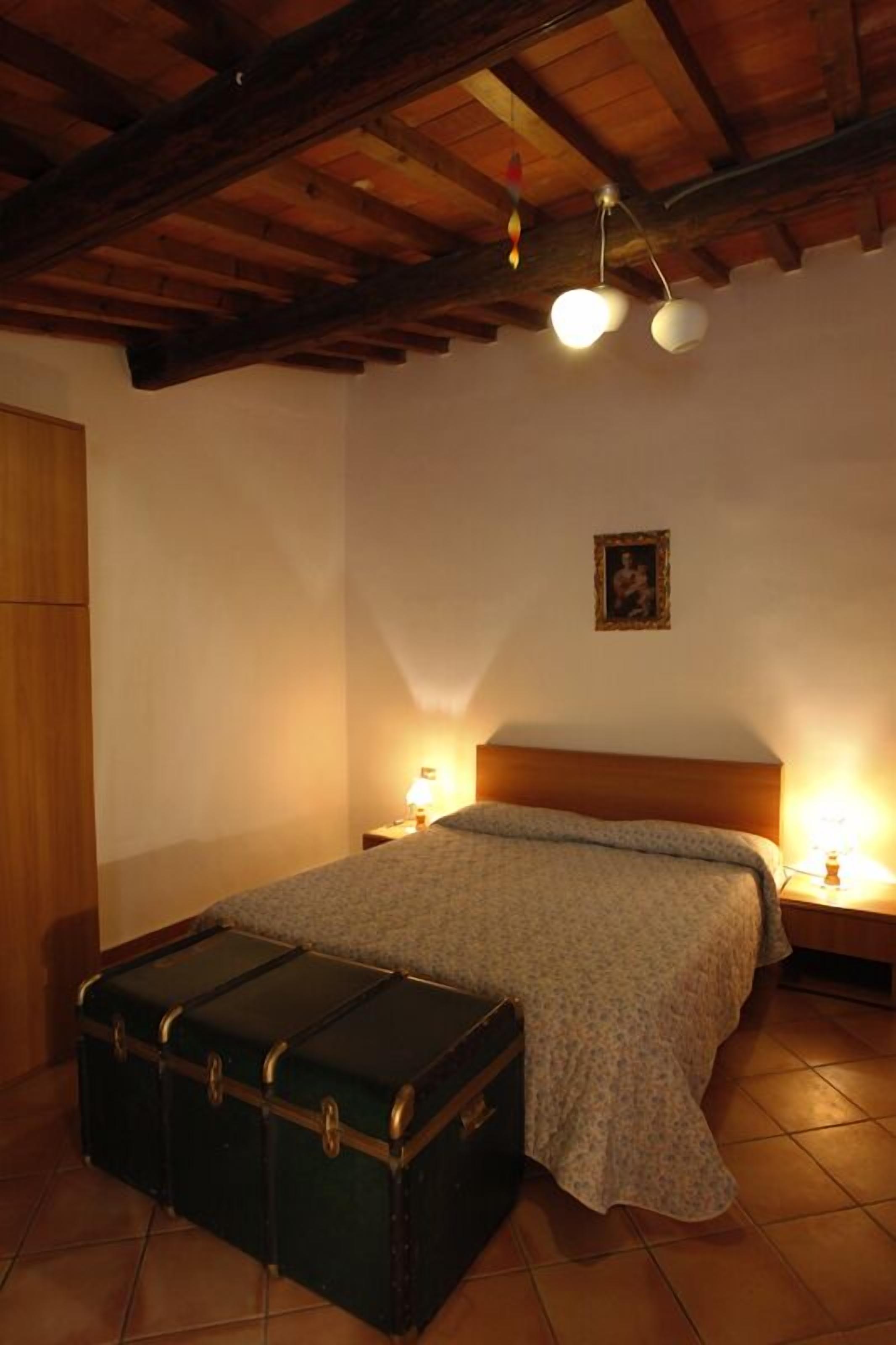 Um quarto com teto de madeira, uma cama com colcha floral, duas luminárias de cabeceira, uma pequena mesa com abajur e uma mala vintage no chão.
