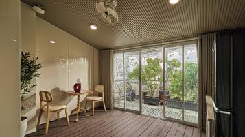 Quarto com piso de madeira, uma mesa, duas cadeiras, um vaso com planta e uma porta de vidro deslizante que dá para área externa com plantas e vista do céu.