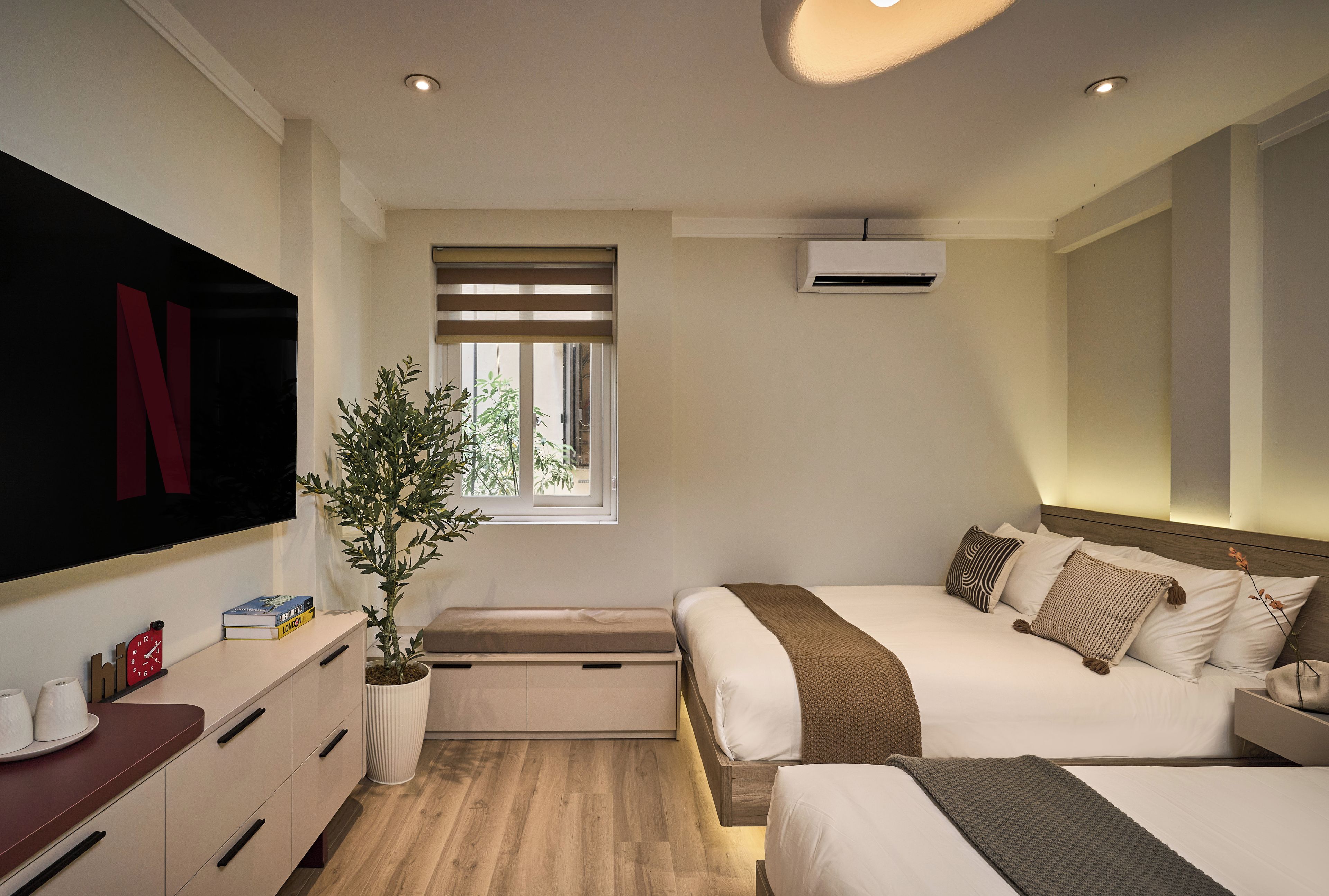 Quarto moderno com televisão, planta em vaso, janela, ar condicionado e cama com travesseiros e manta.