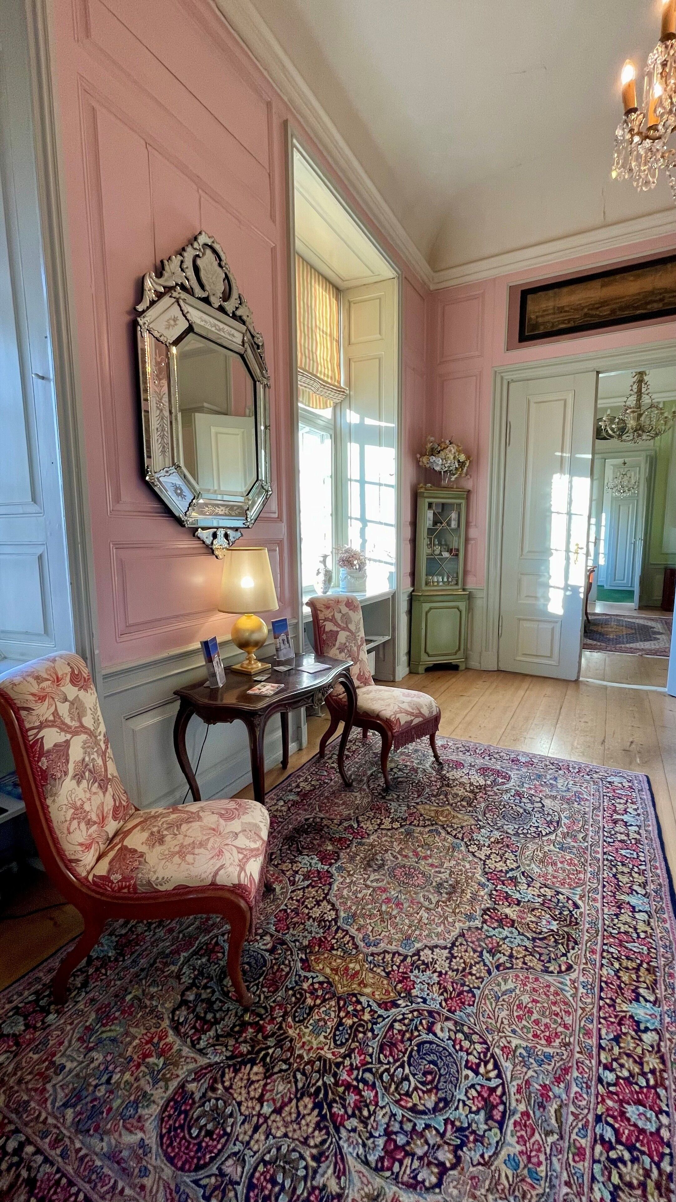 Quarto com paredes rosa, um espelho grande e ornamentado, duas poltronas, um tapete estampado e um candelabro.