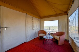 Quarto com tenda, tapete vermelho, duas cadeiras, uma mesa com garrafas de água e vista para a paisagem.