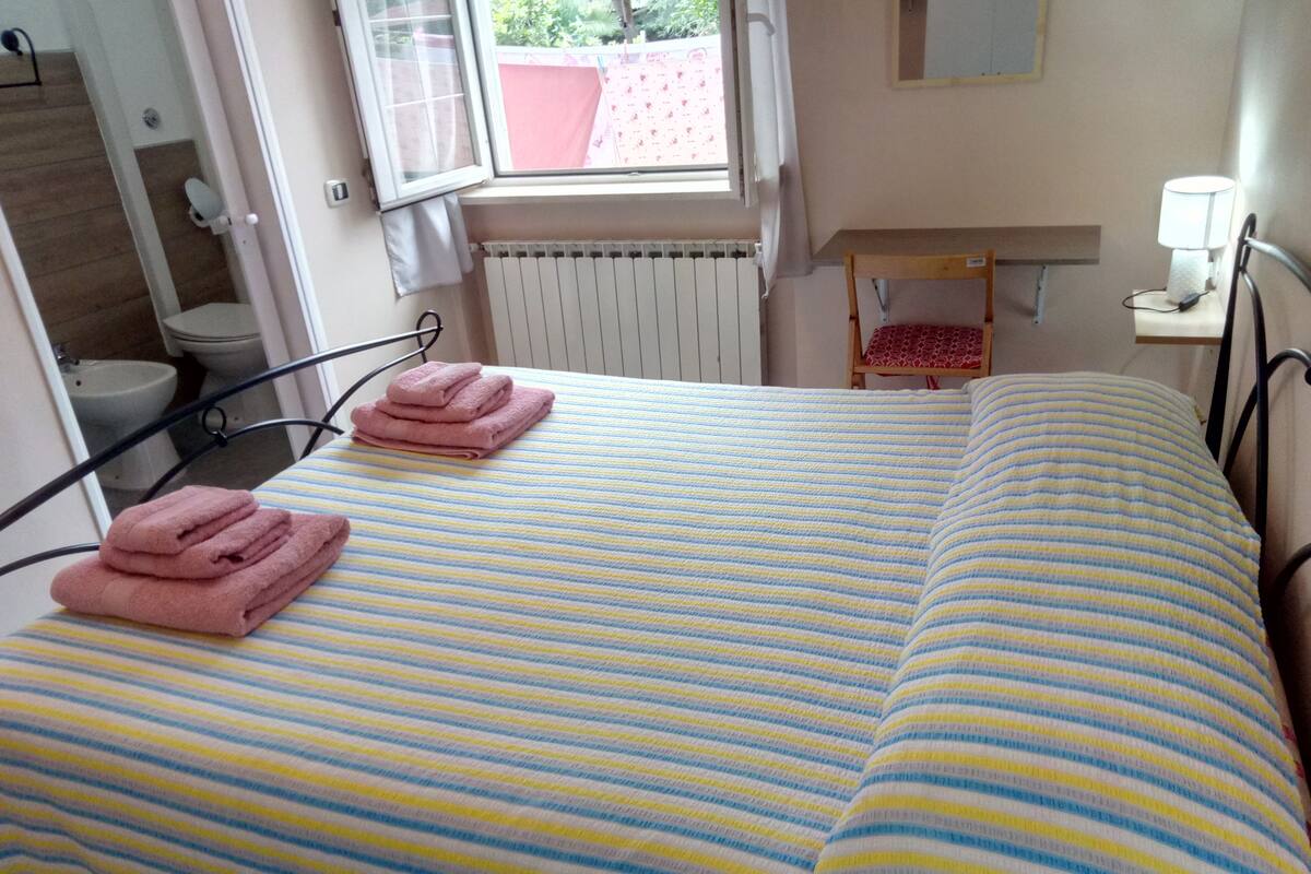 Quarto com cama, janela, radiador, mesa de cabeceira com luminária e vista para o exterior.