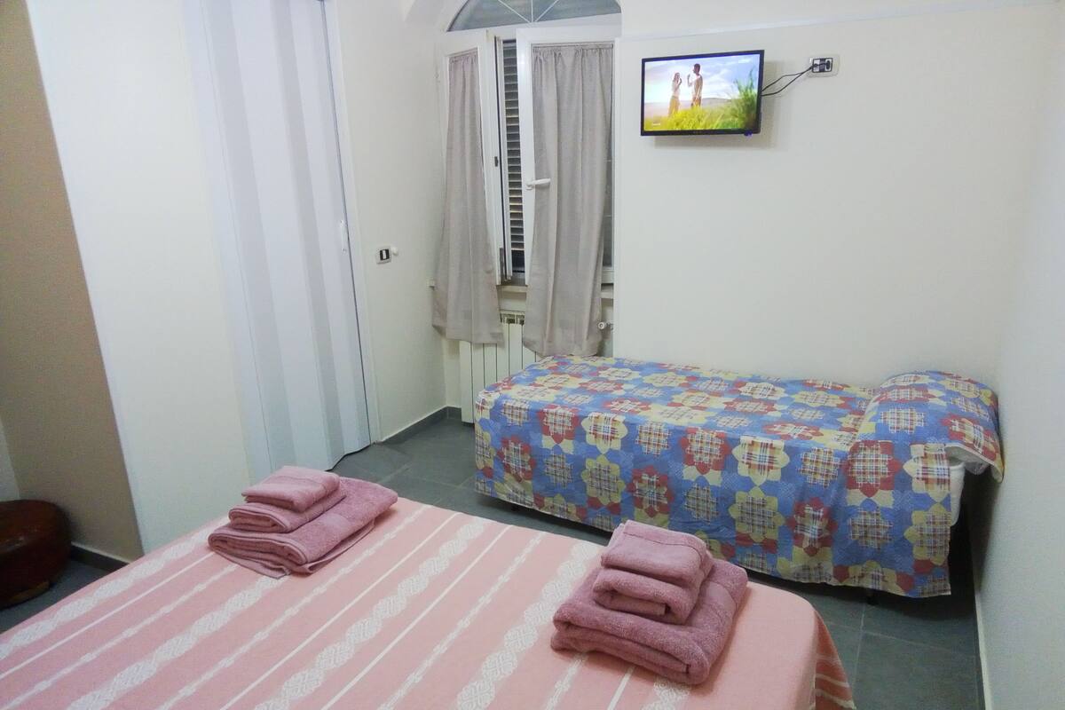 Quarto de hotel com duas camas, cada uma com toalhas cor-de-rosa, uma televisão na parede e uma janela com cortinas.