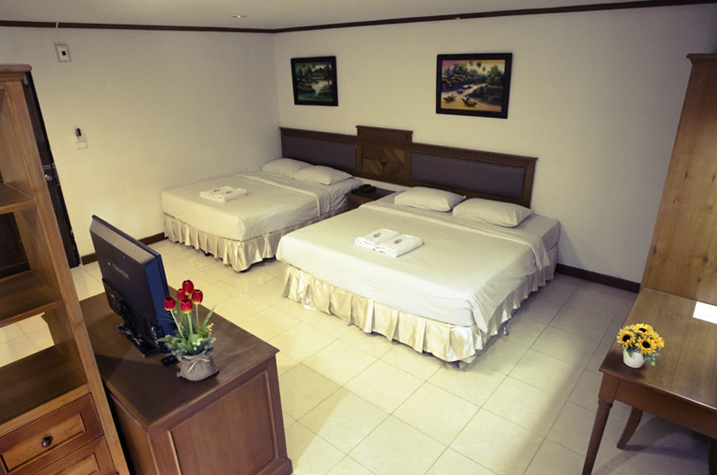 Quarto de hotel com duas camas de solteiro, uma escrivaninha de madeira, televisão e uma pequena mesa com um vaso de flores.