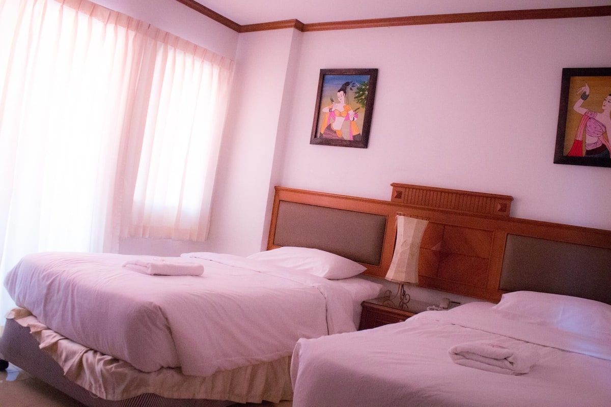 Quarto de hotel com duas camas, cabeceira de madeira, mesa de cabeceira e janela com cortinas leves.
