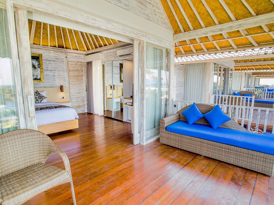 Quarto com piso de madeira, uma cama, um sofá de vime com almofadas azuis e uma cadeira.