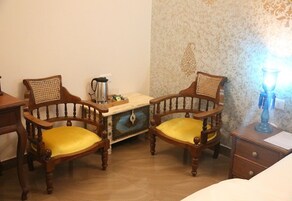 Quarto com duas cadeiras de madeira com almofadas amarelas, uma mesa de madeira pequena com uma chaleira e um abajur em uma mesinha de cabeceira.