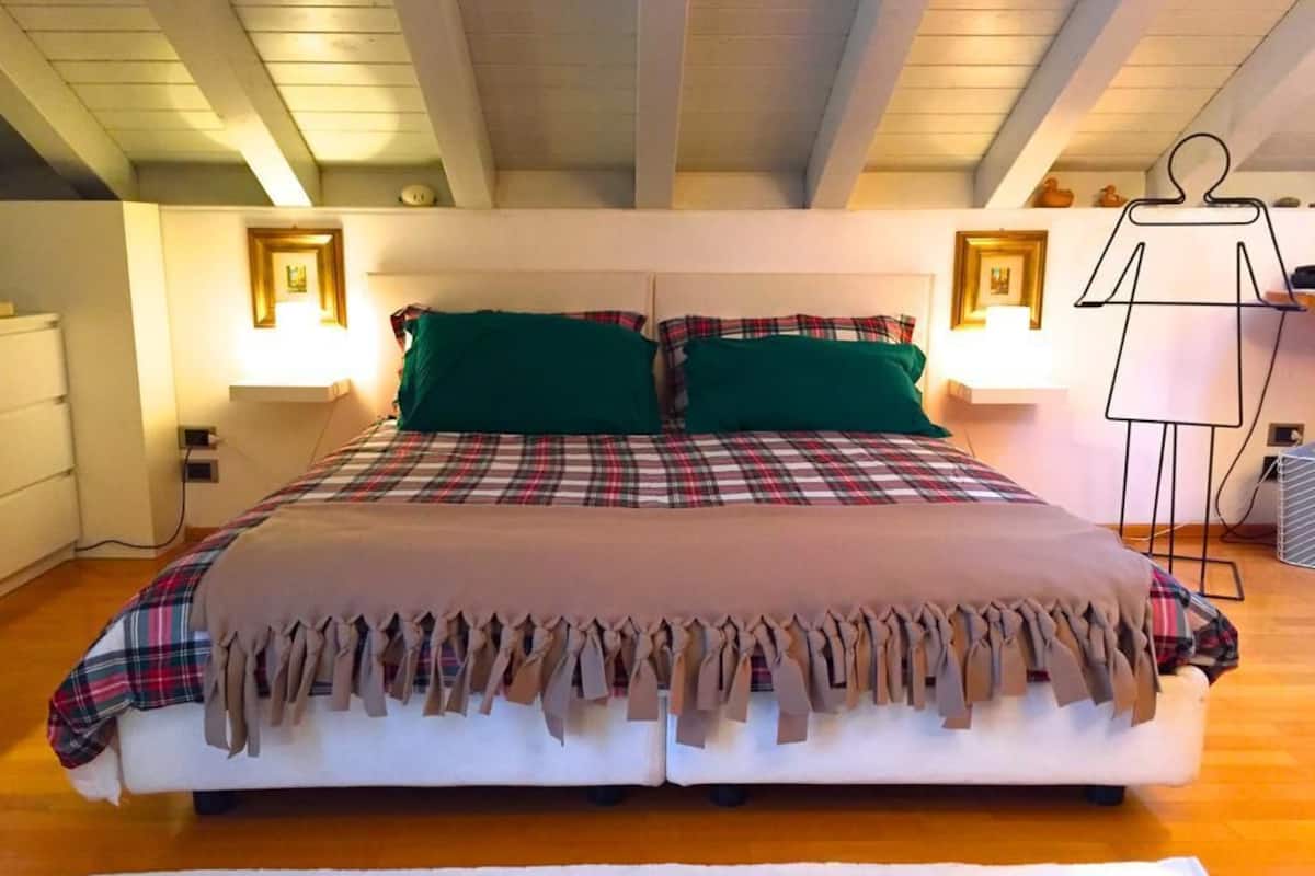 Uma cama de casal com roupa de cama xadrez, duas mesas de cabeceira com luminárias e um teto com vigas aparentes.