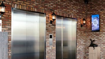 Área de elevadores com paredes de tijolo, duas elevadoras, luminárias fixadas na parede e um visor digital.
