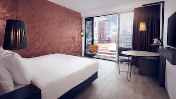 Quarto de hotel com uma cama grande, uma escrivaninha, uma cadeira, uma televisão e vista para a cidade pela janela.