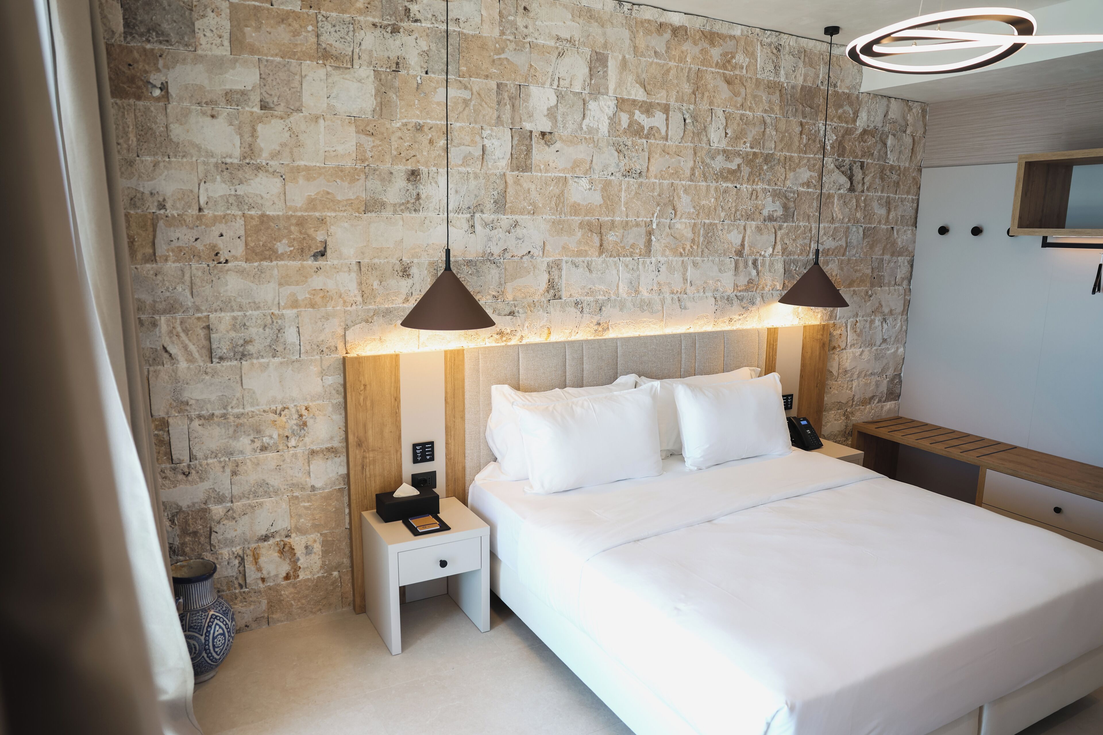Quarto de hotel moderno com parede de pedra, uma cama grande com roupa de cama branca, duas luminárias de cabeceira, uma mesa de cabeceira de madeira e uma pequena mesinha de cabeceira com uma luminária.